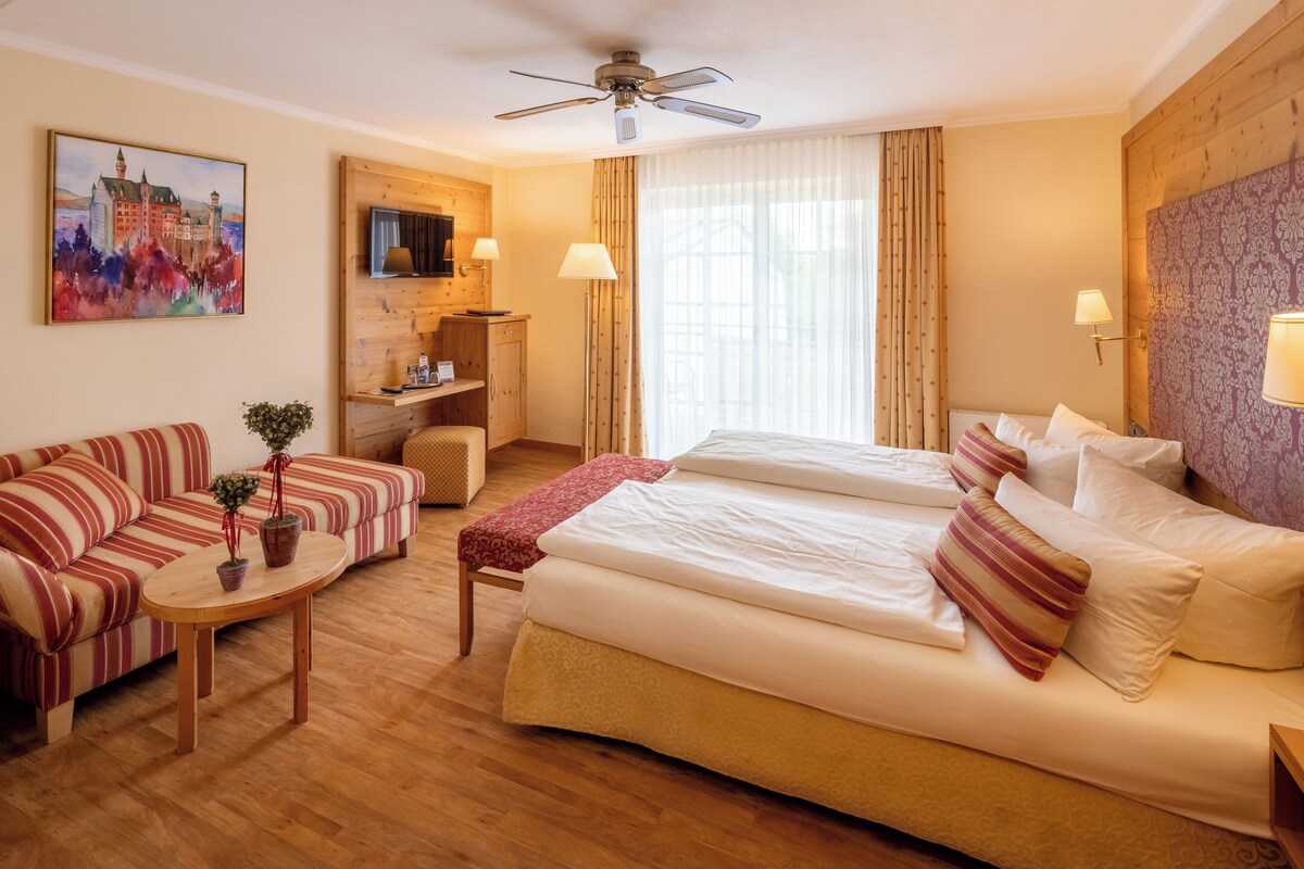 Quarto de hotel com cama, sofá, pequena mesa com planta, ventoinha no teto e um quadro na parede.