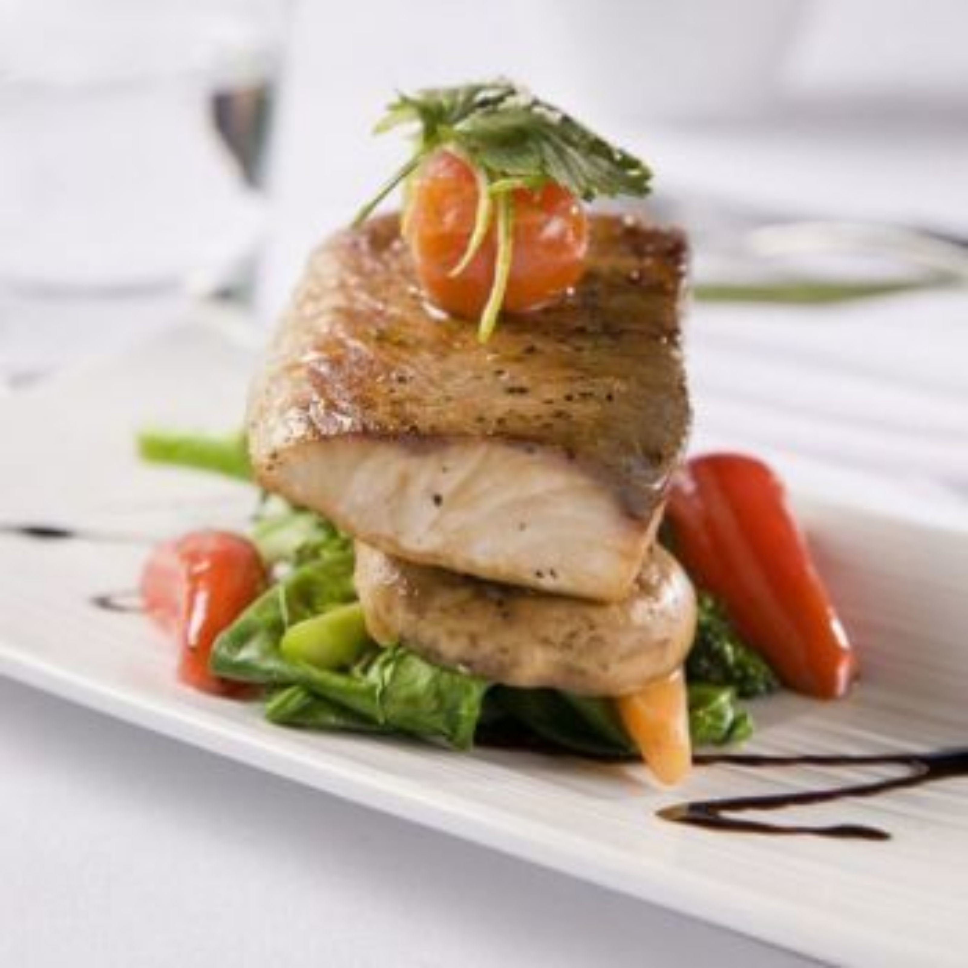 Um prato com um pedaço de peixe grelhado, adornado com um tomate cereja e um ramo de salsa, acompanhado de um guarnição de vegetais e uma fatia de cenoura.