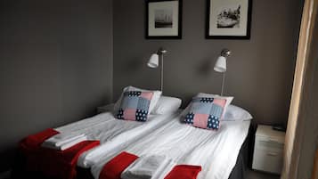 Uma cama de casal com roupa de cama branca e detalhes vermelhos, duas luminárias de cabeceira, um criado-mudo com um rádio e duas fotos emolduradas na parede.
