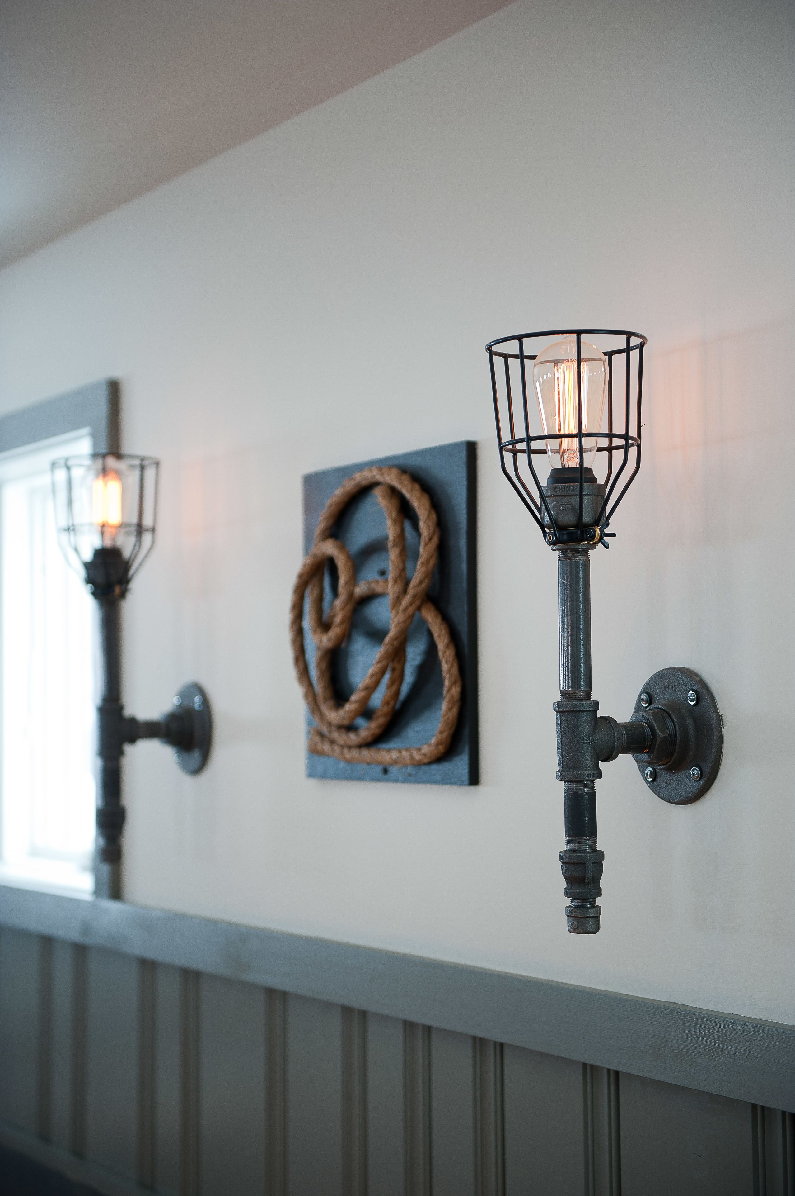 Duas luminárias fixadas na parede com lâmpadas aparentes, uma obra de arte com corda e uma parede branca com uma janela.