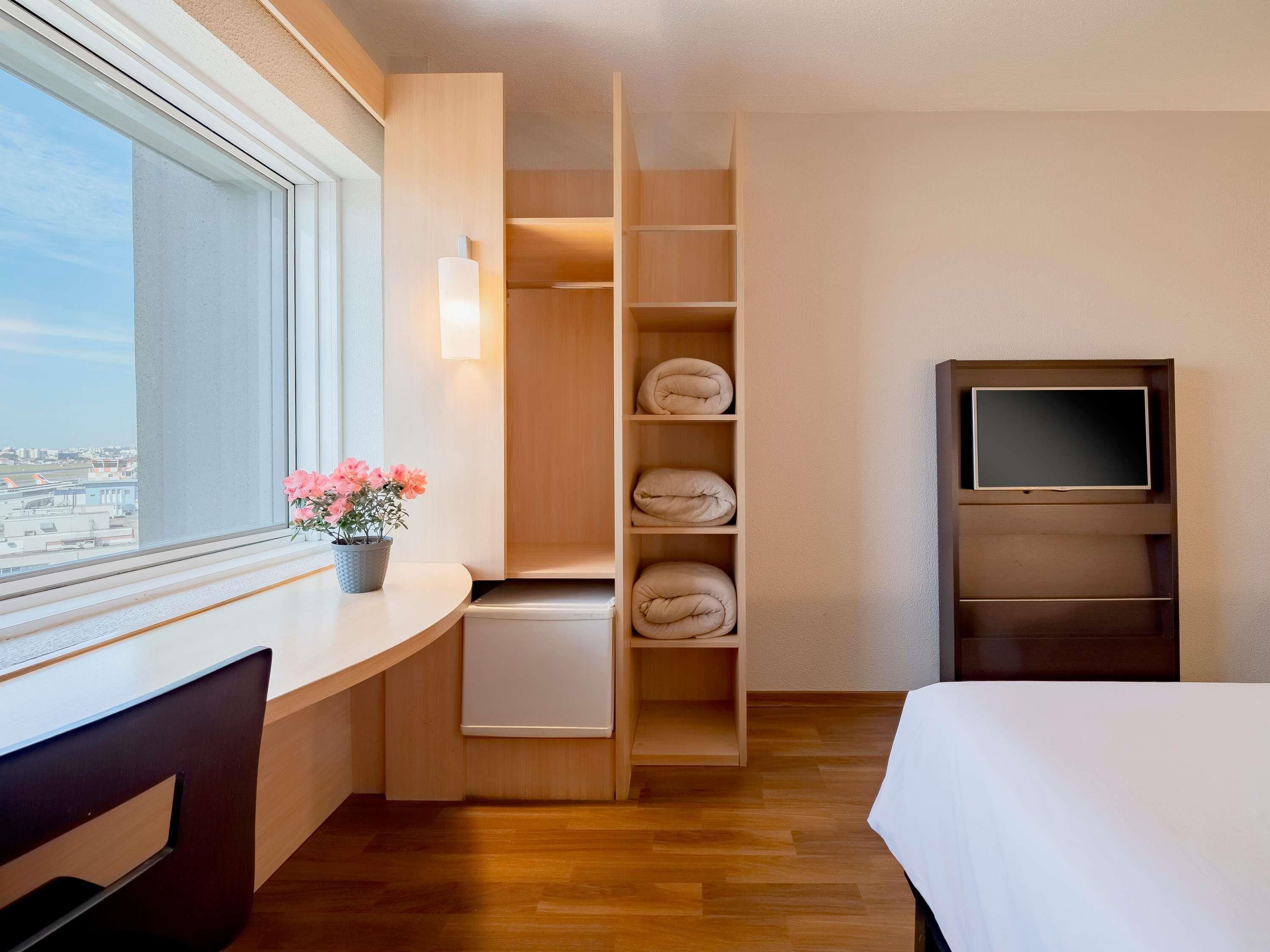 Quarto de hotel com cama, televisão, secretária com cadeira, janela com vista para o aeroporto e prateleira embutida com toalhas.