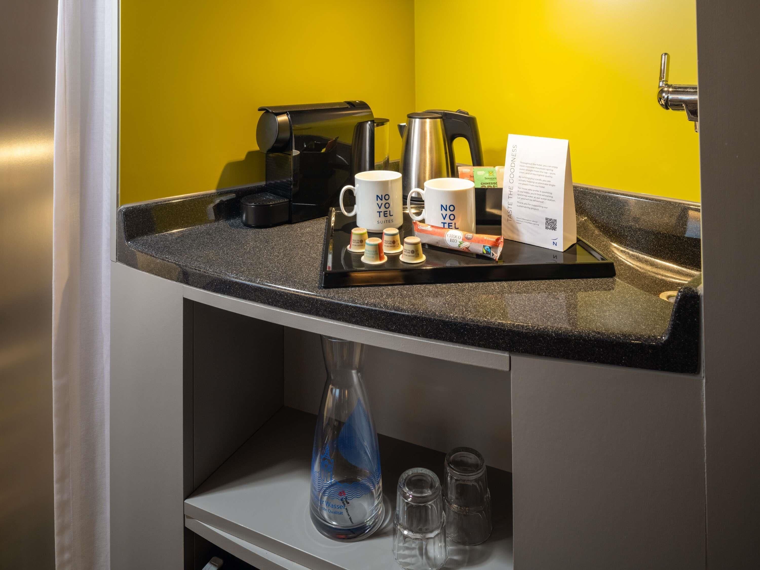 Cozinha compacta de hotel com cafeteira, chaleira e bandeja com xícaras de café e acompanhamentos.