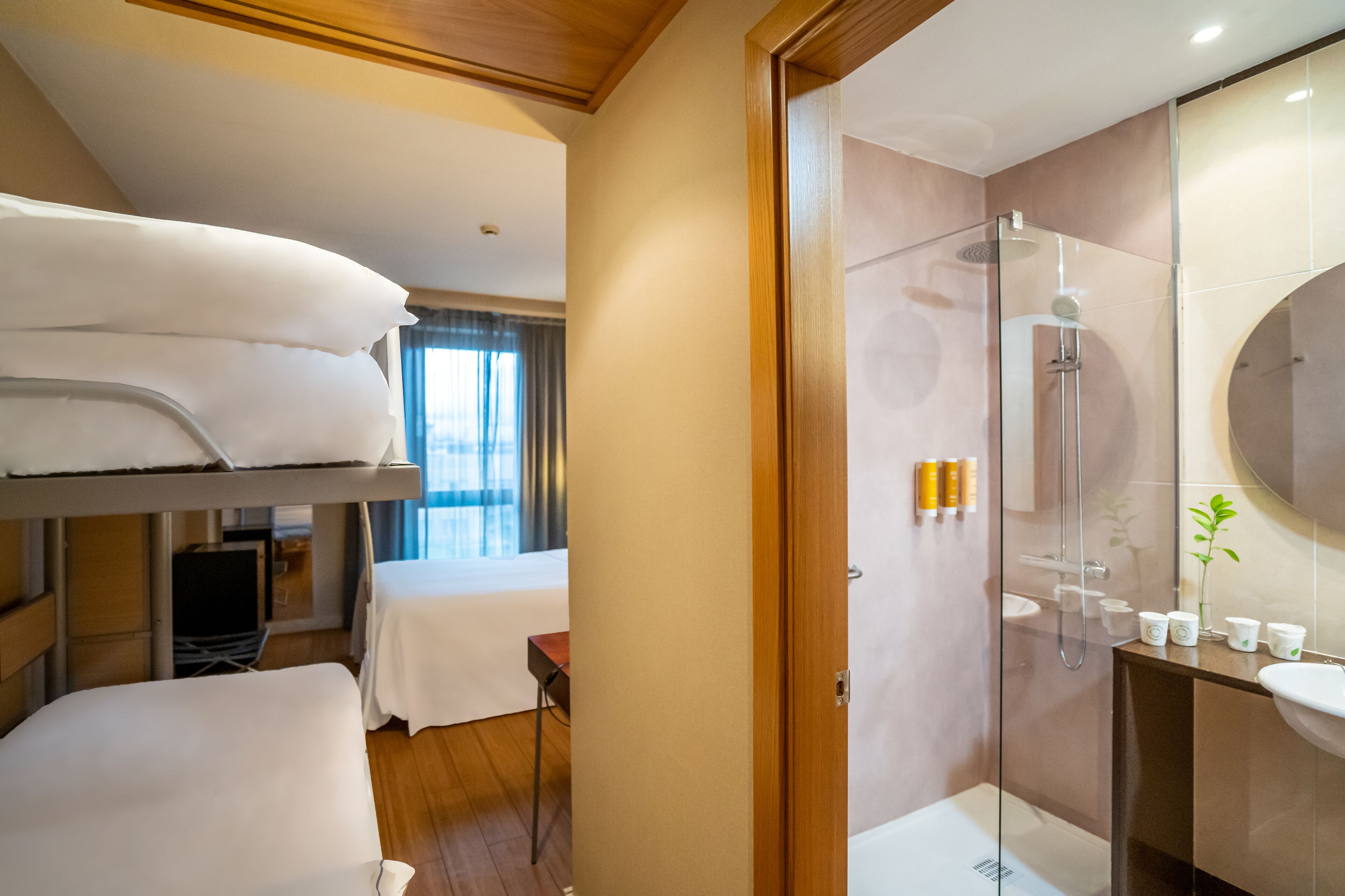 Quarto de hotel com beliches, chuveiro, pia e artigos de higiene no balcão.