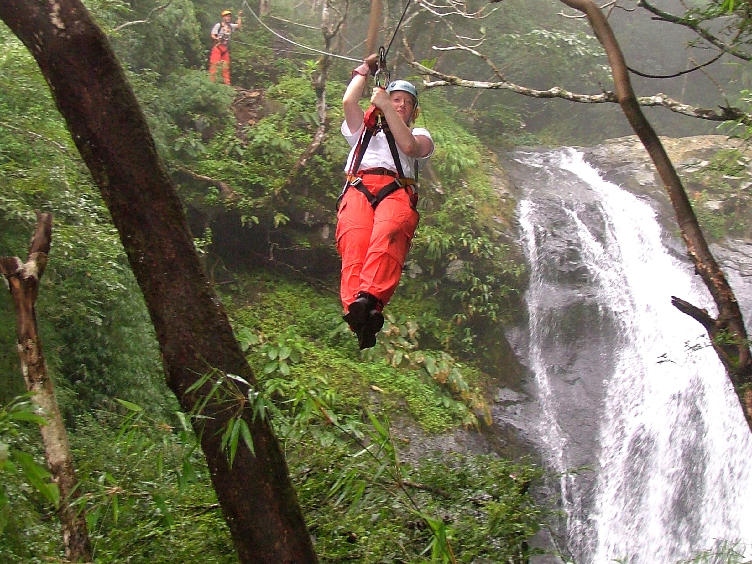 Uma pessoa praticando tirolesa em meio a uma floresta, com uma cachoeira ao fundo.