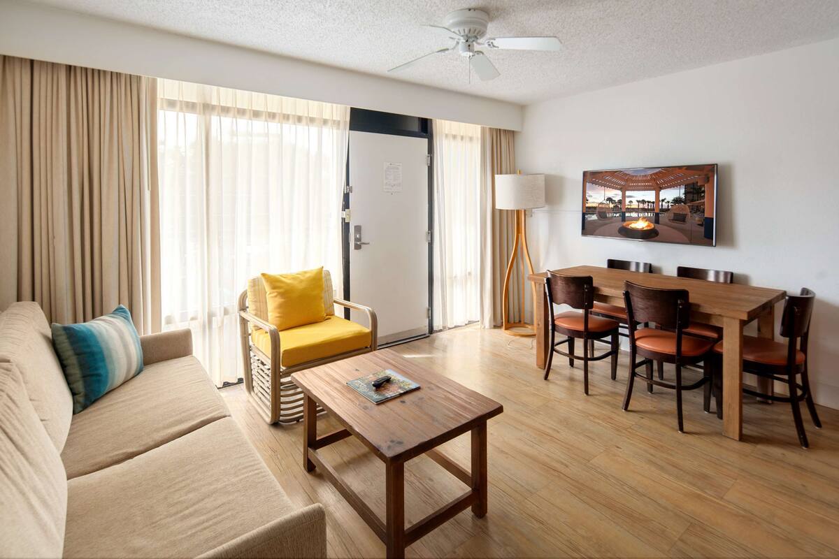 Uma sala de estar moderna com sofá, poltrona amarela, mesa de centro de madeira, mesa de jantar com cadeiras e um ventilador de teto.