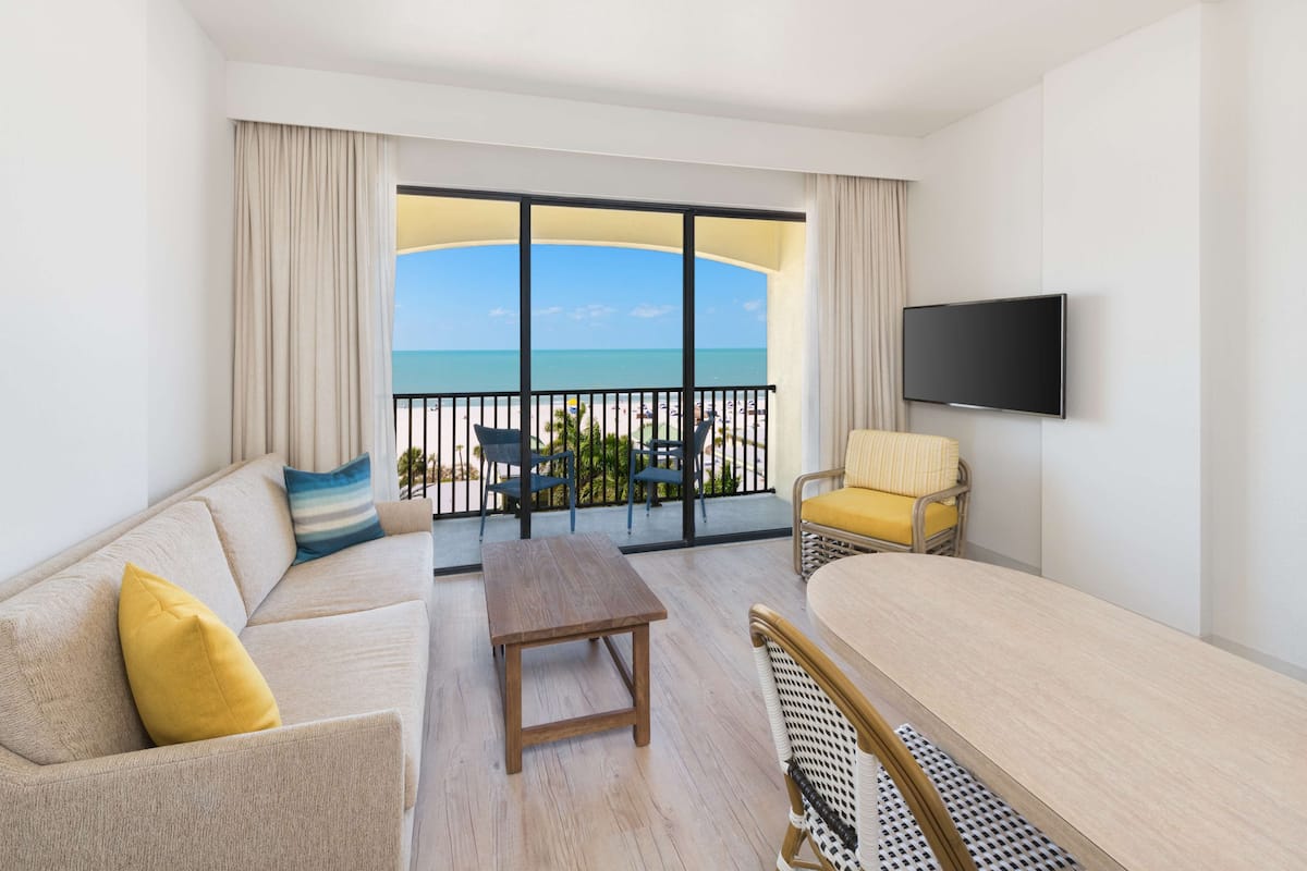Quarto de hotel com varanda, um sofá, uma cadeira, uma mesa pequena e uma televisão.