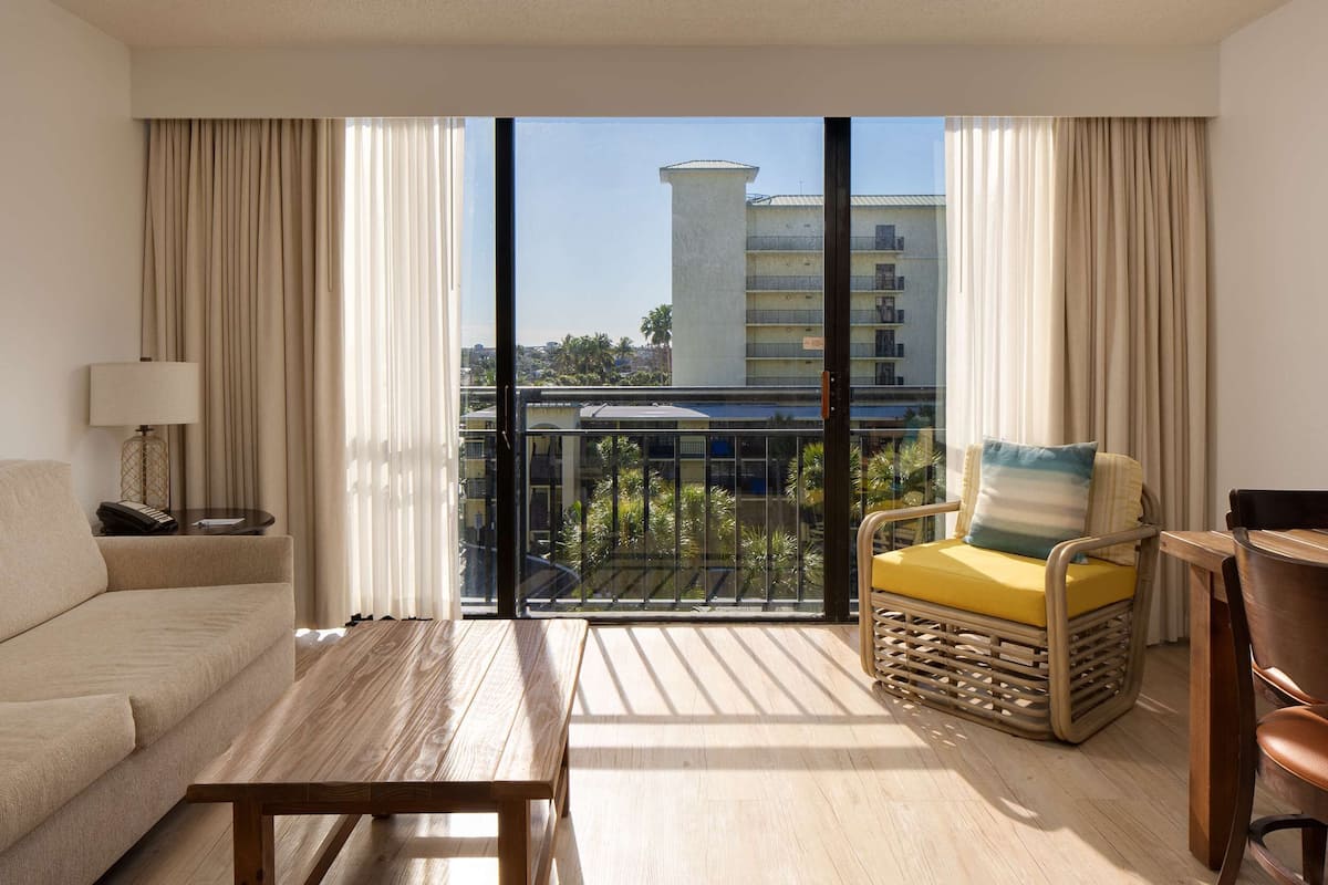 Quarto de hotel com varanda, sofá, poltrona, mesa e luminária.