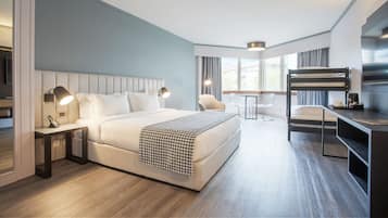 Quarto de hotel moderno com uma cama grande, mesa de cabeceira com abajur, escrivaninha com cadeira e vista da cidade através de amplas janelas.