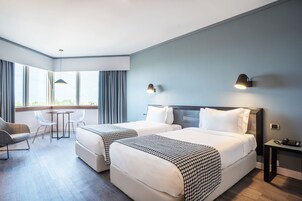 Quarto de hotel com duas camas, uma escrivaninha, uma cadeira e vista para o exterior.