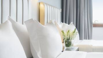 Quarto de hotel com cama bem arrumada, mesa de cabeceira com um vaso de flores, e vista do céu pelas cortinas.