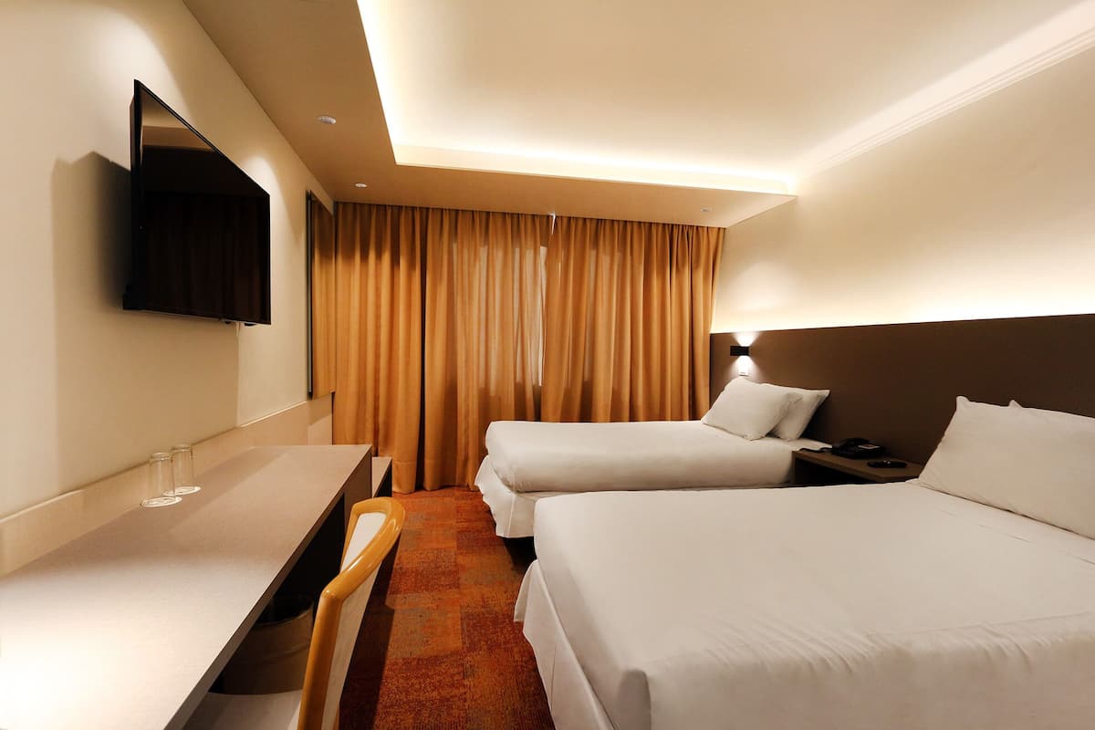 Quarto de hotel com duas camas, uma escrivaninha, uma cadeira, televisão na parede e janela com cortinas.