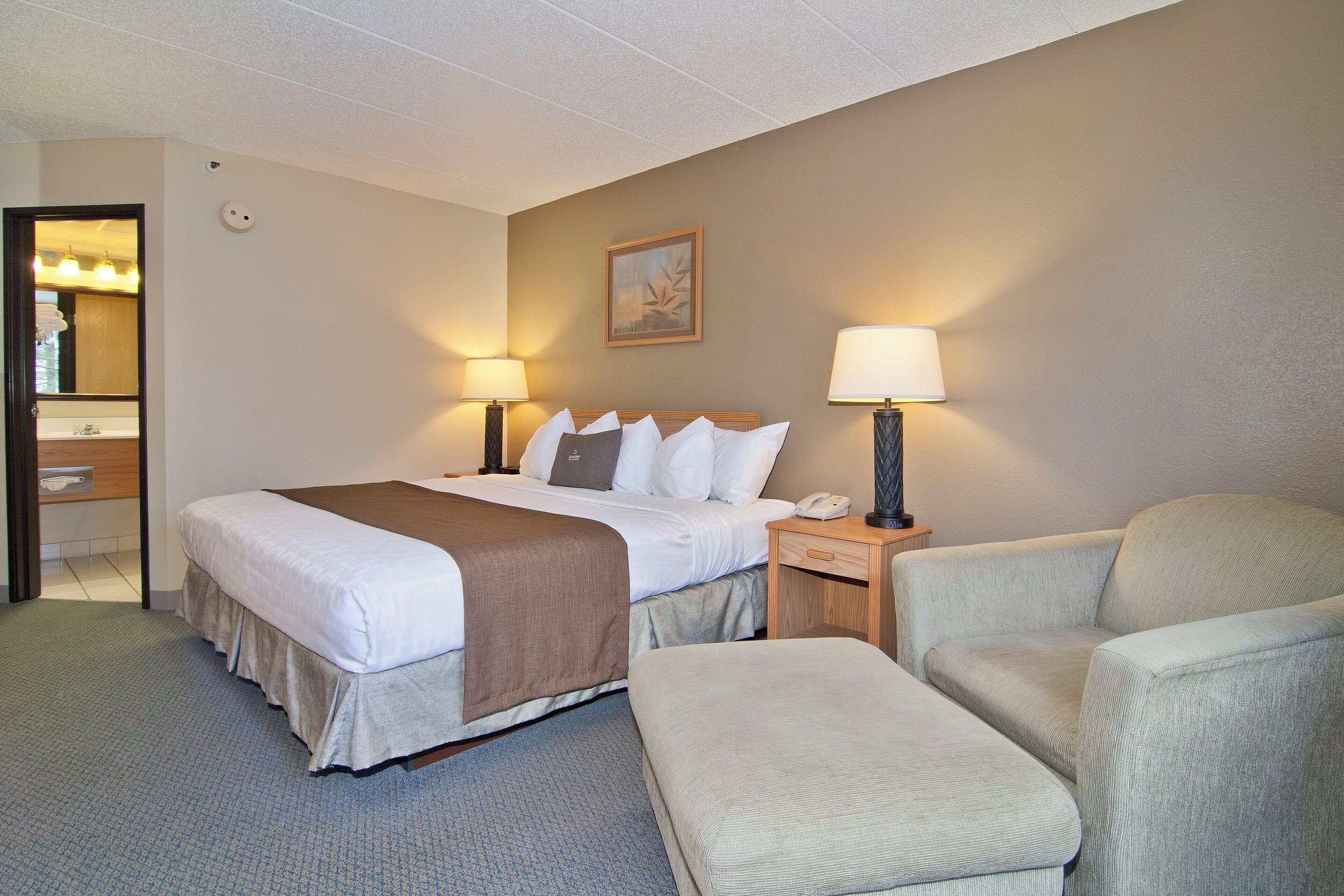 Quarto de hotel com cama, mesa de cabeceira, luminária, cadeira e uma pequena mesa com telefone.