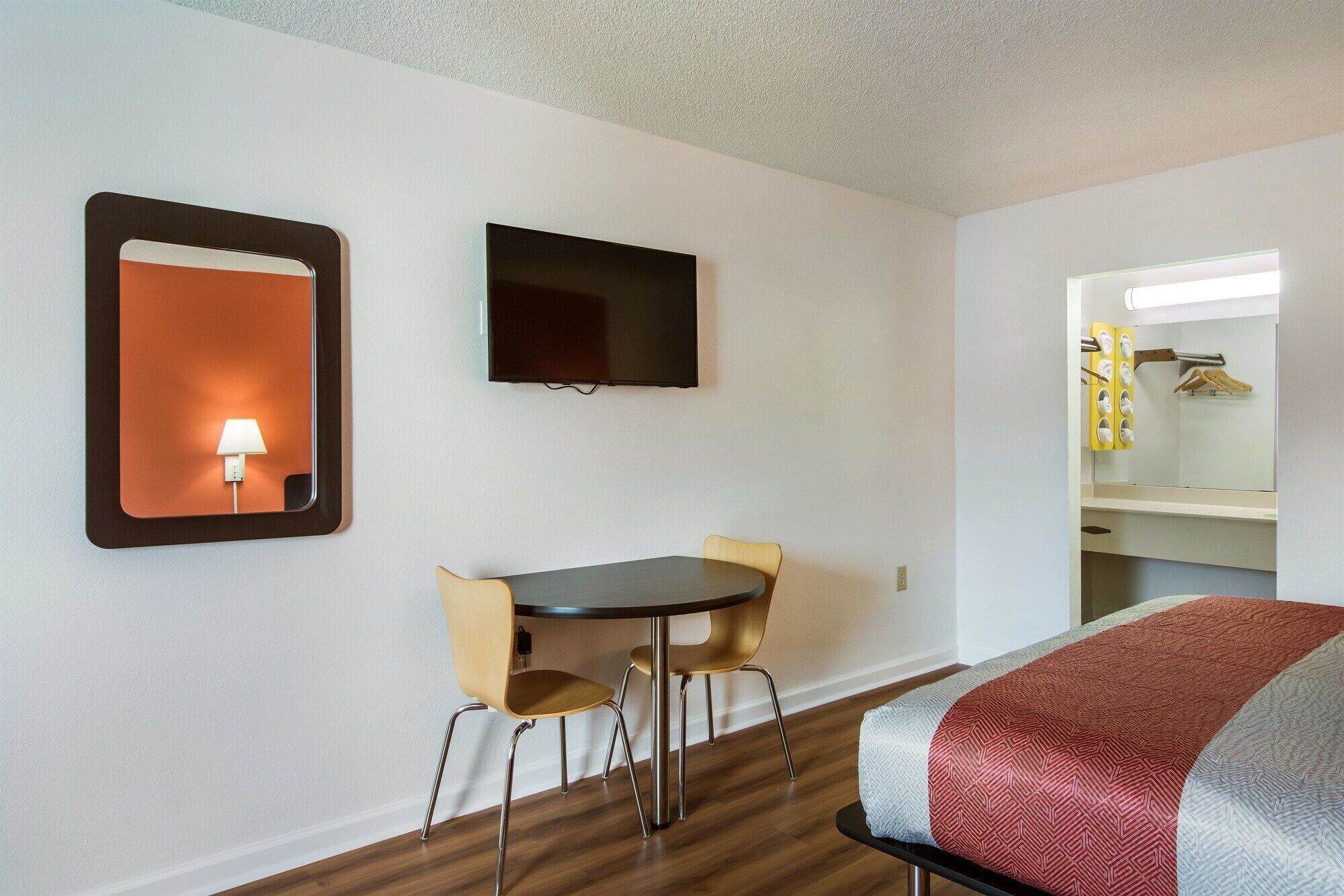 Quarto de hotel com uma cama, uma mesa com duas cadeiras, uma TV fixada na parede e um espelho com uma luminária.