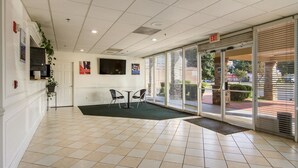 Um hall limpo e bem iluminado, com piso de azulejos, balcão de recepção, duas cadeiras, uma televisão e uma janela grande com persianas.