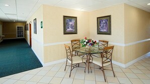 Quarto de hotel com uma mesa de vidro redonda, quatro cadeiras de madeira e um arranjo floral.