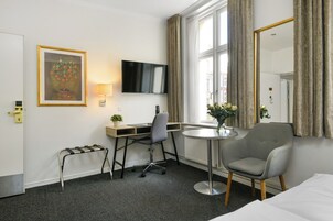 Quarto de hotel com cama, escrivaninha com cadeira, televisão, mesa pequena com vaso e janela com cortinas.