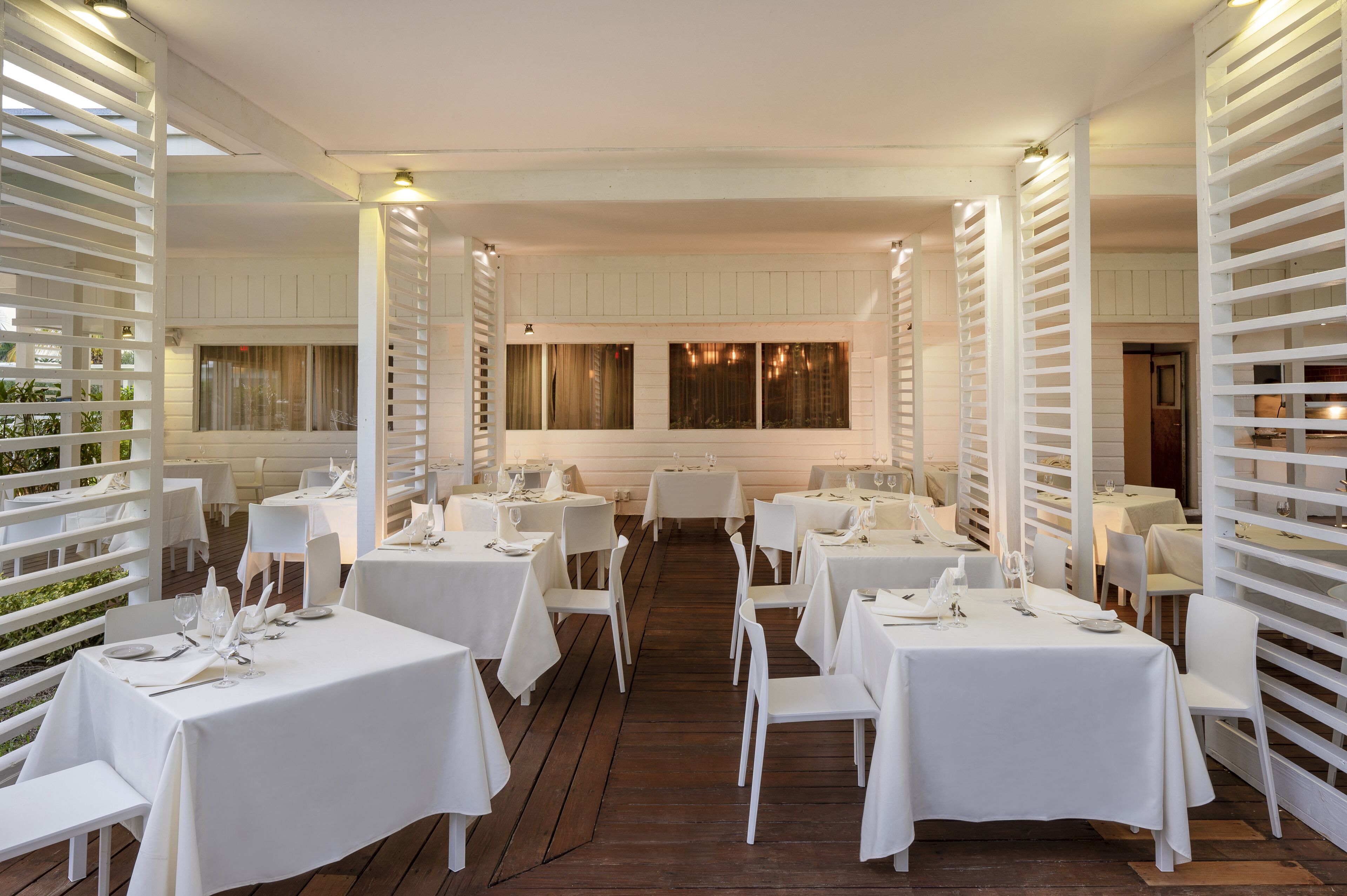 Restaurante com mesas e cadeiras brancas, piso de madeira e paredes brancas com ripas verticais.