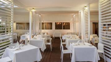 Restaurante com mesas e cadeiras brancas, piso de madeira e paredes brancas com ripas verticais.
