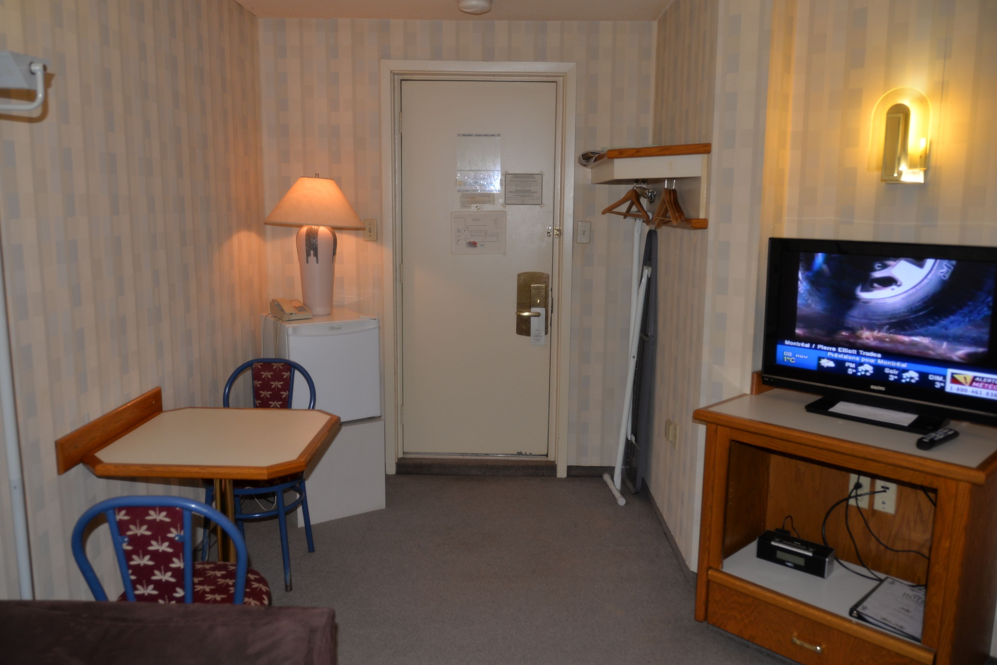 Quarto de hotel com televisão, uma mesinha e duas cadeiras, um abajur e um porta-roupas.