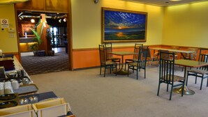 Restaurante com pintura de pôr do sol, várias mesas e cadeiras, e um buffet com diversos pratos e acompanhamentos.