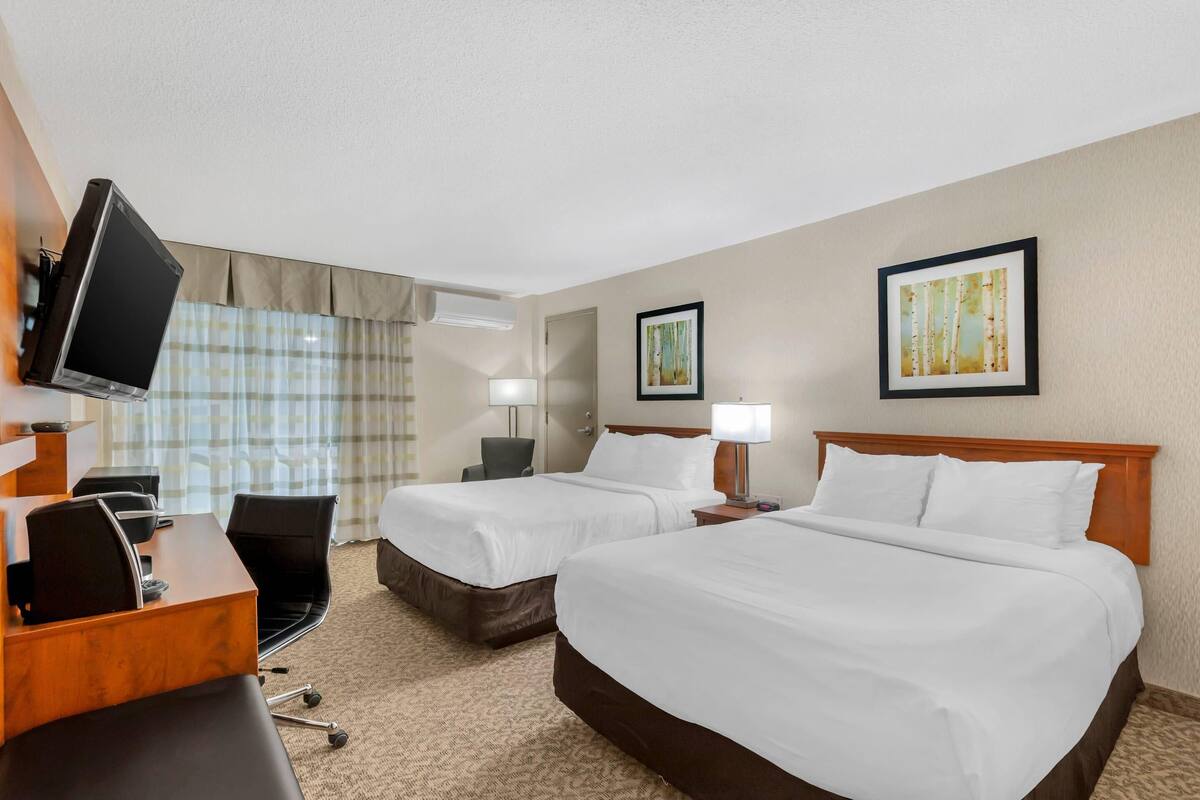 Quarto de hotel com duas camas, uma escrivaninha com computador, uma cadeira e uma televisão.