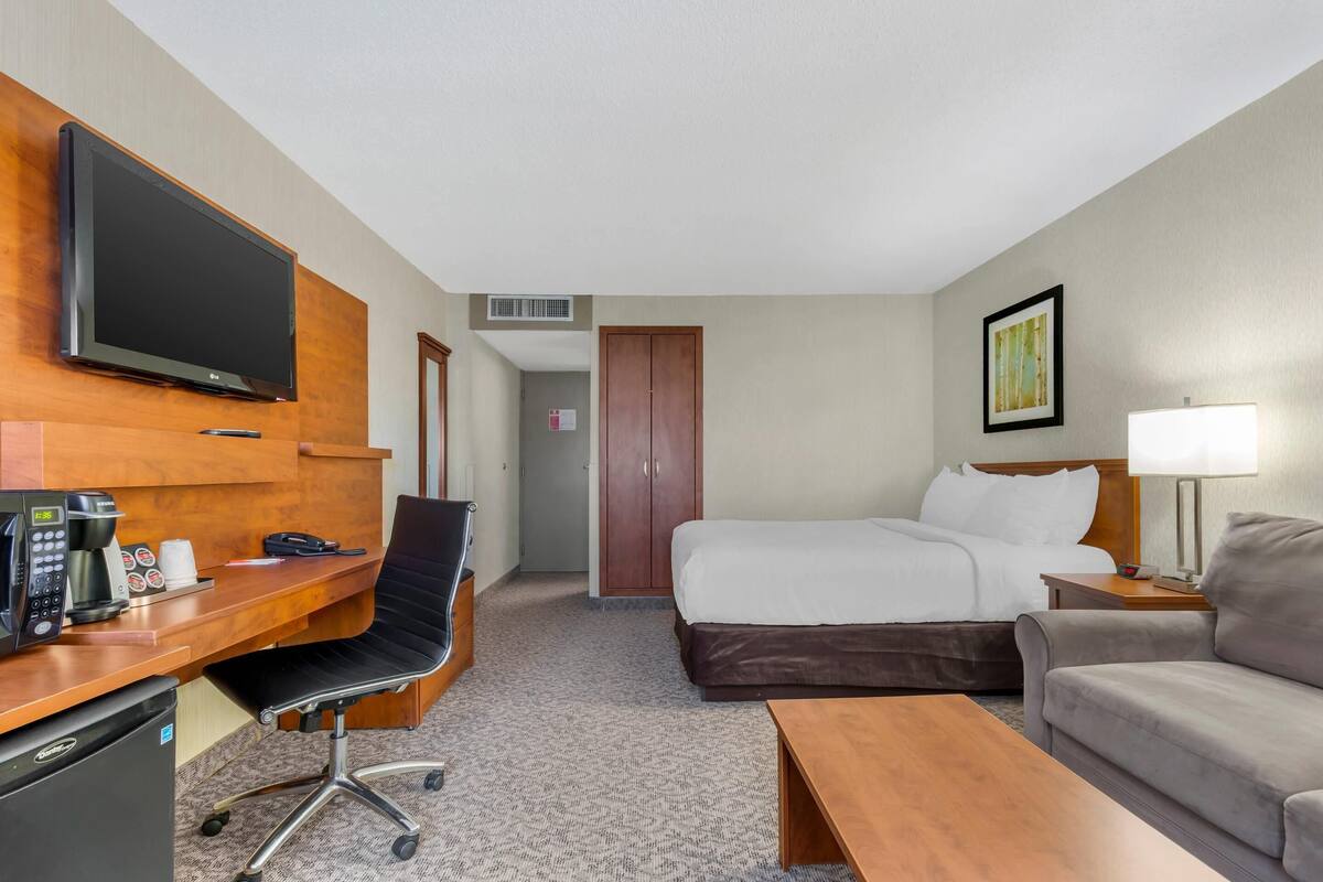 Quarto de hotel com cama, escrivaninha com computador, cadeira, televisão, cafeteira e luminária.