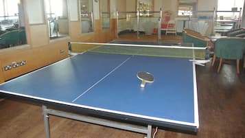 Uma mesa de pingue-pongue em um espaço com piso de madeira, cadeiras verdes e uma área de recreação infantil ao fundo.