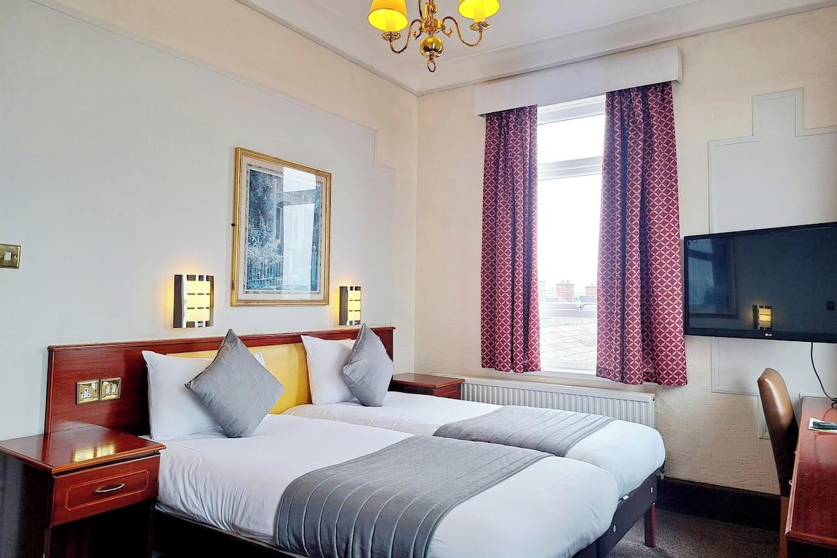 Quarto de hotel com cama de casal, mesas de cabeceira, televisão, janela com cortinas, lustre e um quadro emoldurado na parede.