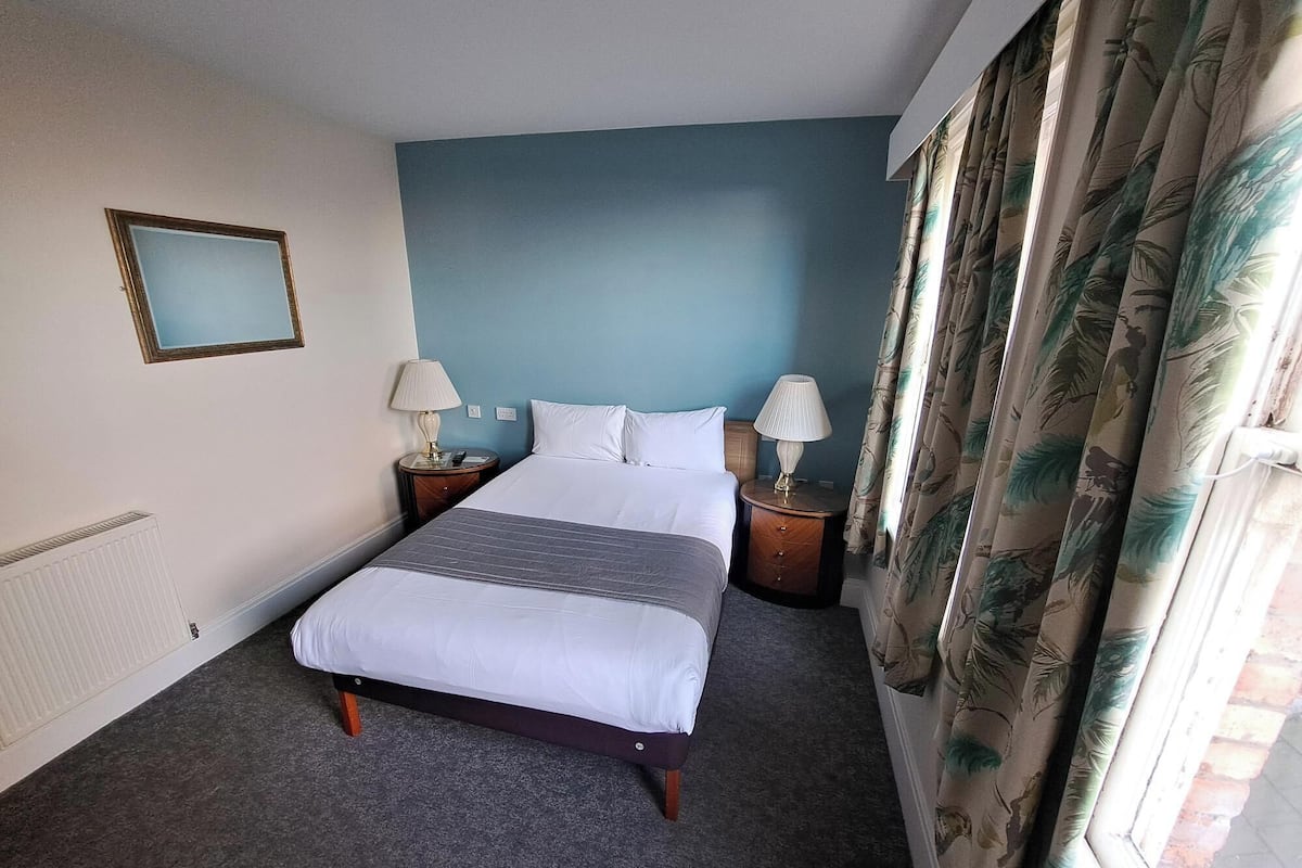 Quarto de hotel com uma cama grande, duas mesas de cabeceira com luminárias, um espelho na parede e uma janela com cortinas.