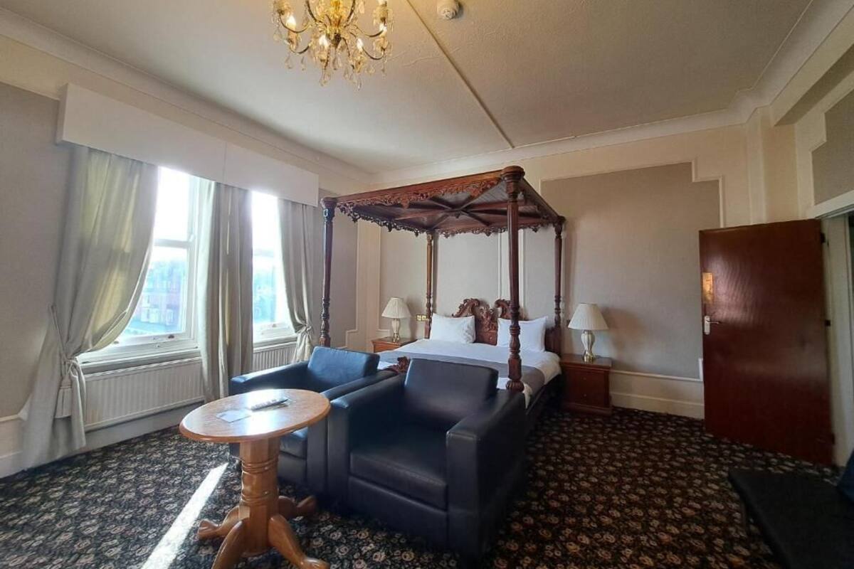 Quarto de hotel com cama de dossel, área de estar com mesa e cadeiras, um lustre e uma grande janela com cortinas.