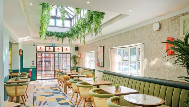 Restaurante com assentos em verde, mesas de madeira e plantas penduradas.