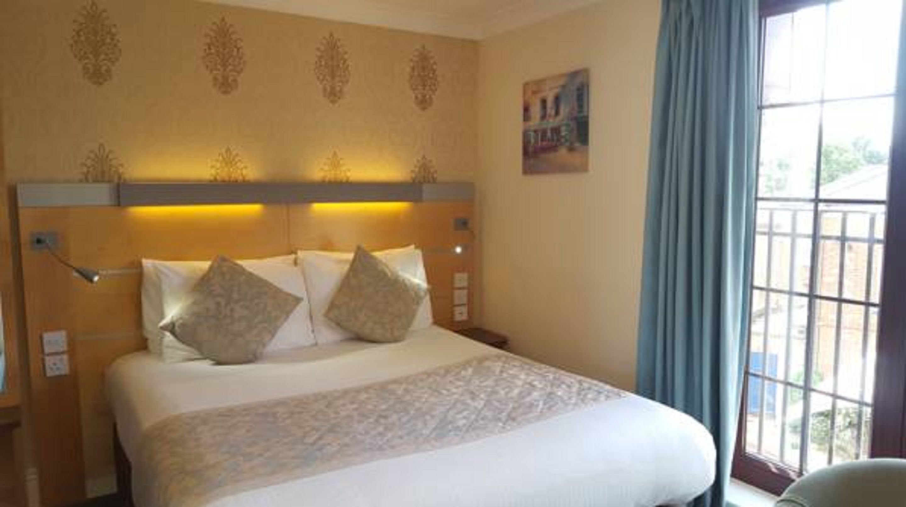 Quarto de hotel com uma cama grande, cabeceira com iluminação embutida, um quadro na parede e uma janela com cortinas.