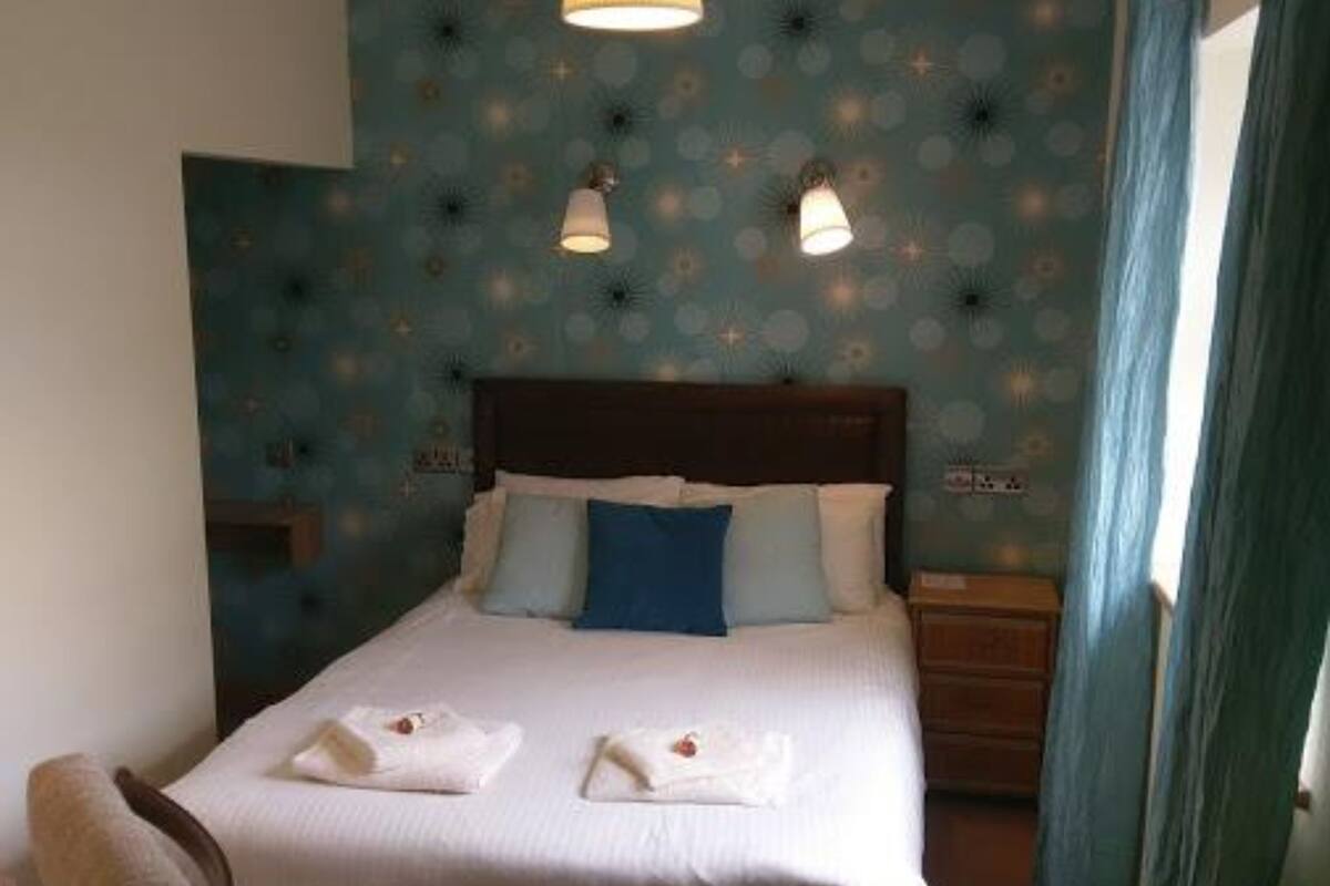 Cama bem arrumada com lençóis brancos e duas almofadas, cabeceira de madeira e papel de parede azul com padrão.