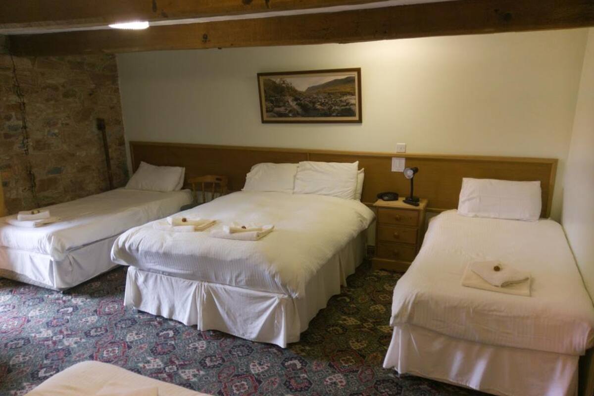 Quarto de hotel com três camas de solteiro, cabeceira de madeira, mesinha de cabeceira com abajur e piso coberto por carpete.