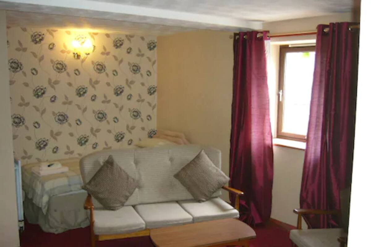 Sala de estar com sofá, uma mesinha e uma cadeira. Há uma janela com cortinas e uma parede com padrão floral.