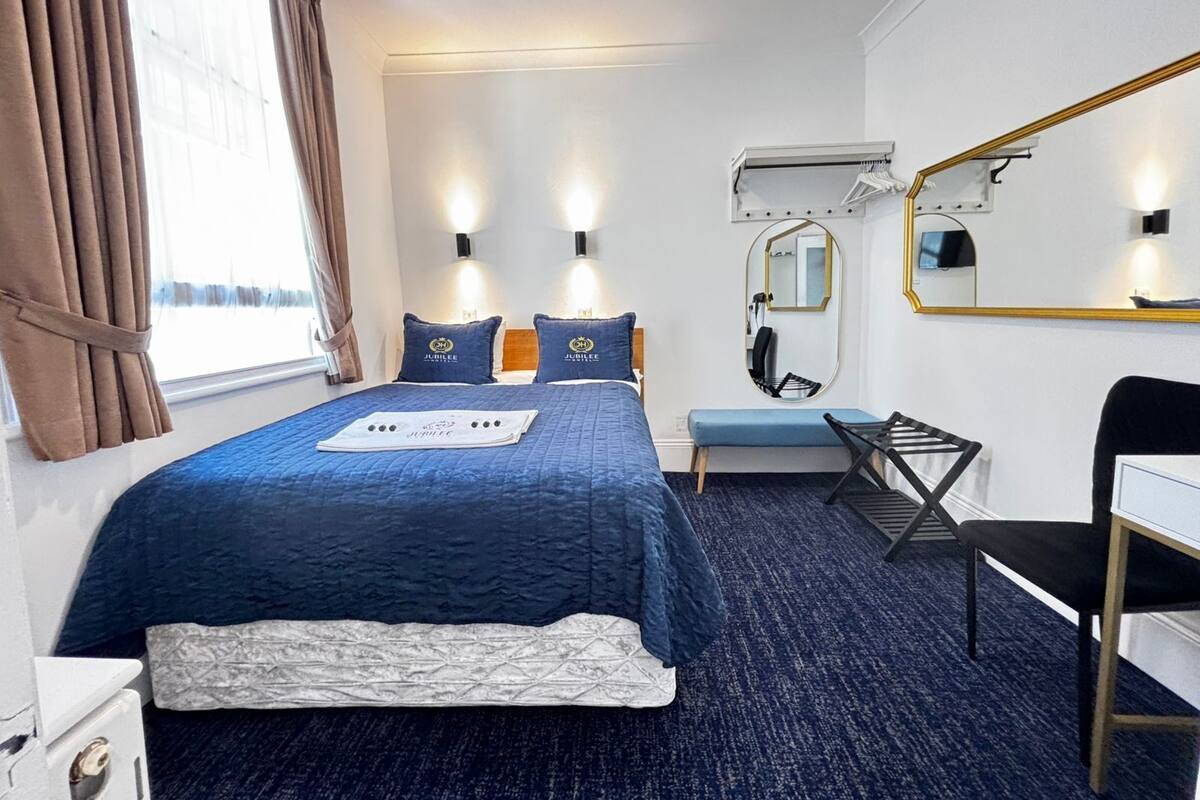 Quarto de hotel com cama, escrivaninha, cadeira, espelho e janela com cortinas.