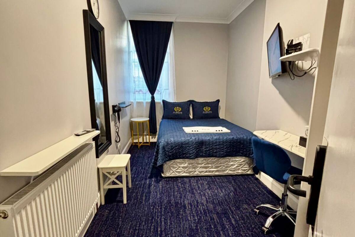 Quarto com cama, escrivaninha, cadeira, radiador, espelho e televisão na parede.