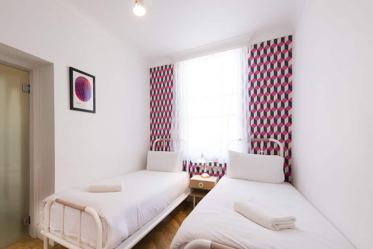 Quarto pequeno e limpo com duas camas de solteiro, mesa de cabeceira com abajur e janela com cortinas estampadas.