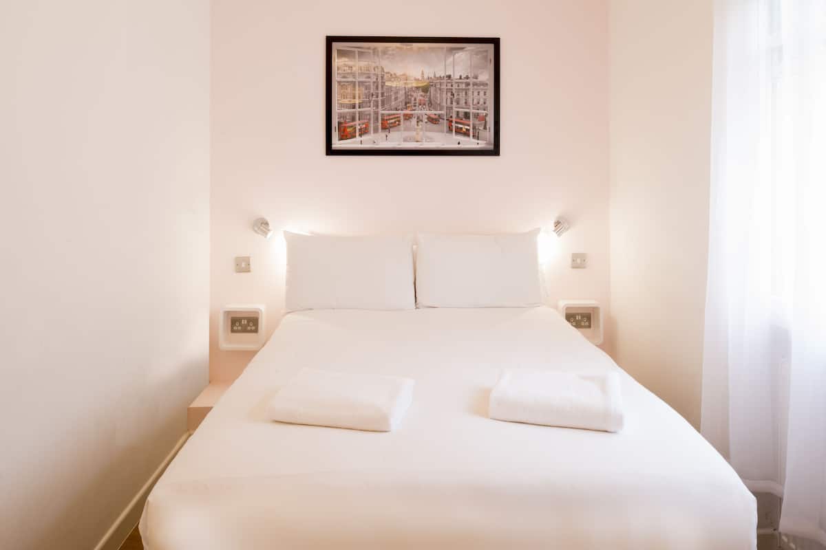 Cama bem arrumada com lençóis brancos e duas almofadas, uma obra de arte com paisagem urbana emoldurada acima da cama, e abajures de cada lado.