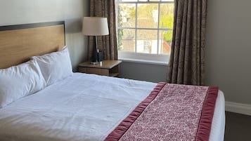 Cama bem arrumada com roupa de cama branca e vermelha, um abajur e uma janela com cortinas estampadas.
