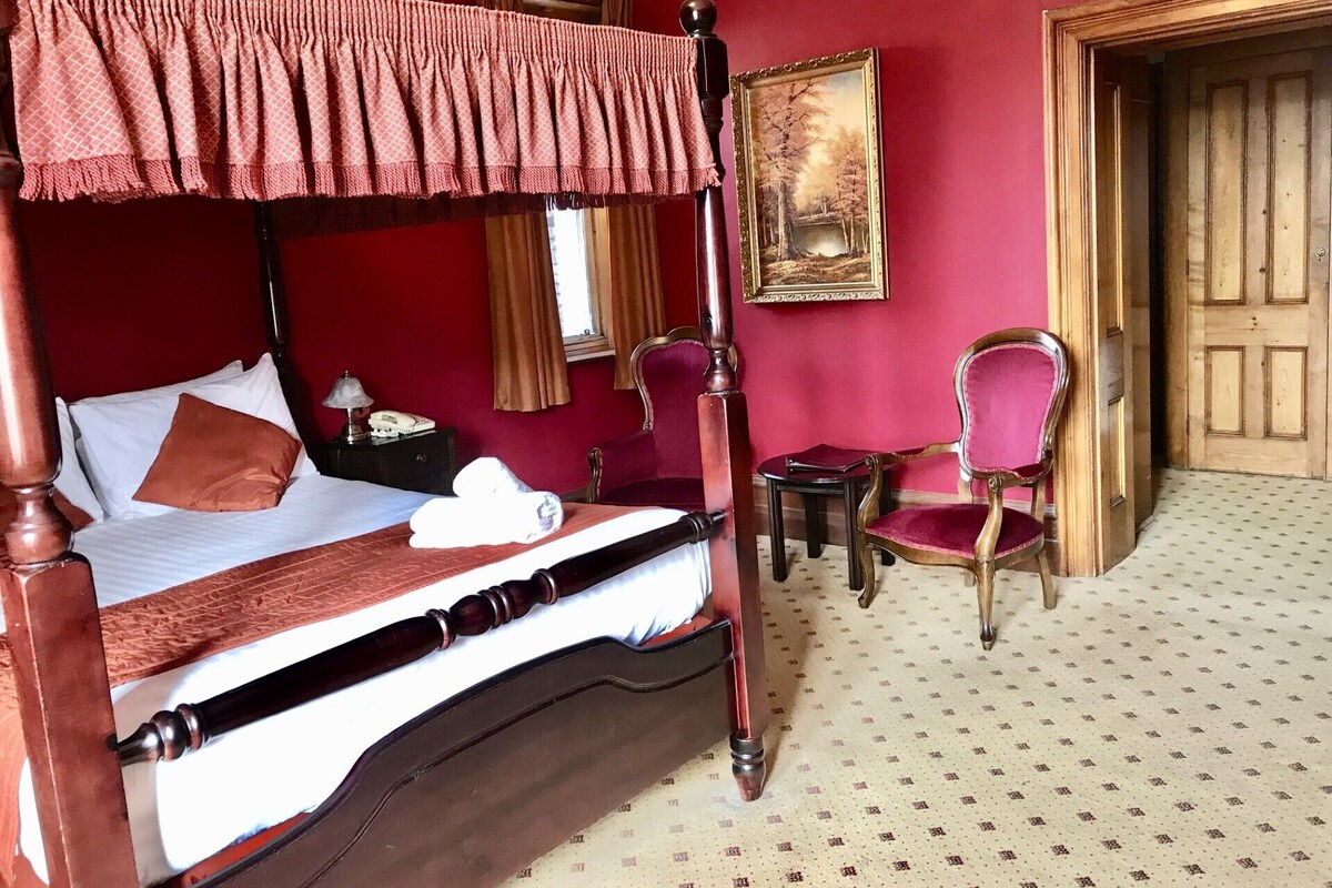 Uma cama de dossel com dossel vermelho, uma mesa de cabeceira de madeira com uma luminária, uma cadeira e um quadro na parede.