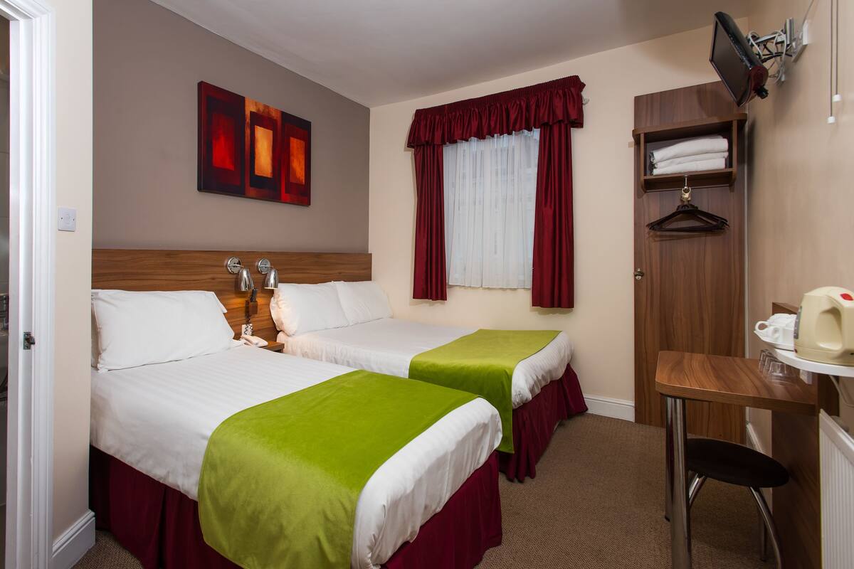 Quarto de hotel com duas camas, uma escrivaninha, uma cadeira, uma pia e uma janela com cortinas.