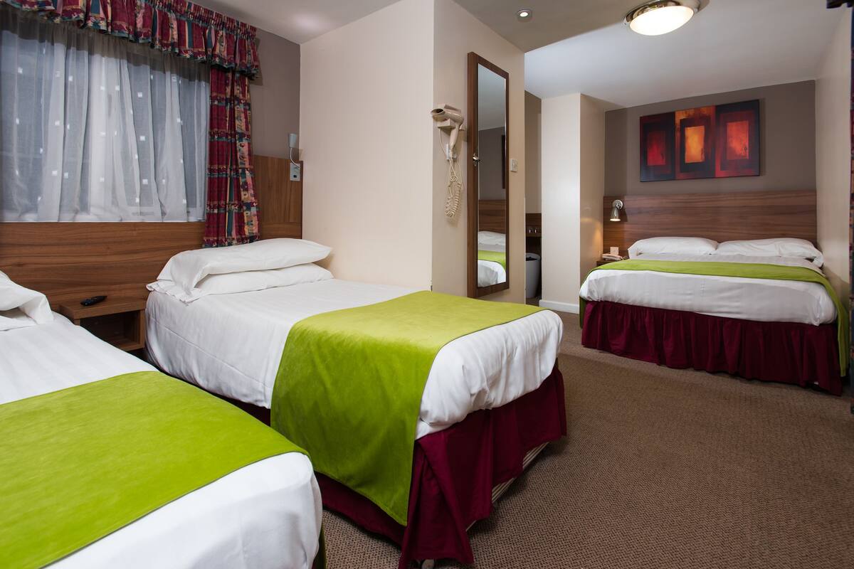 Quarto de hotel com duas camas, cada uma com colcha verde e branca, cabeceira de madeira, espelho, mesa de cabeceira com abajur e janela com cortinas finas.