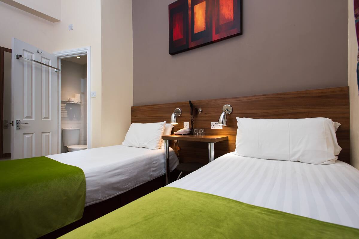 Quarto de hotel com duas camas, cabeceira de madeira, mesa de cabeceira com abajur e um quadro na parede.