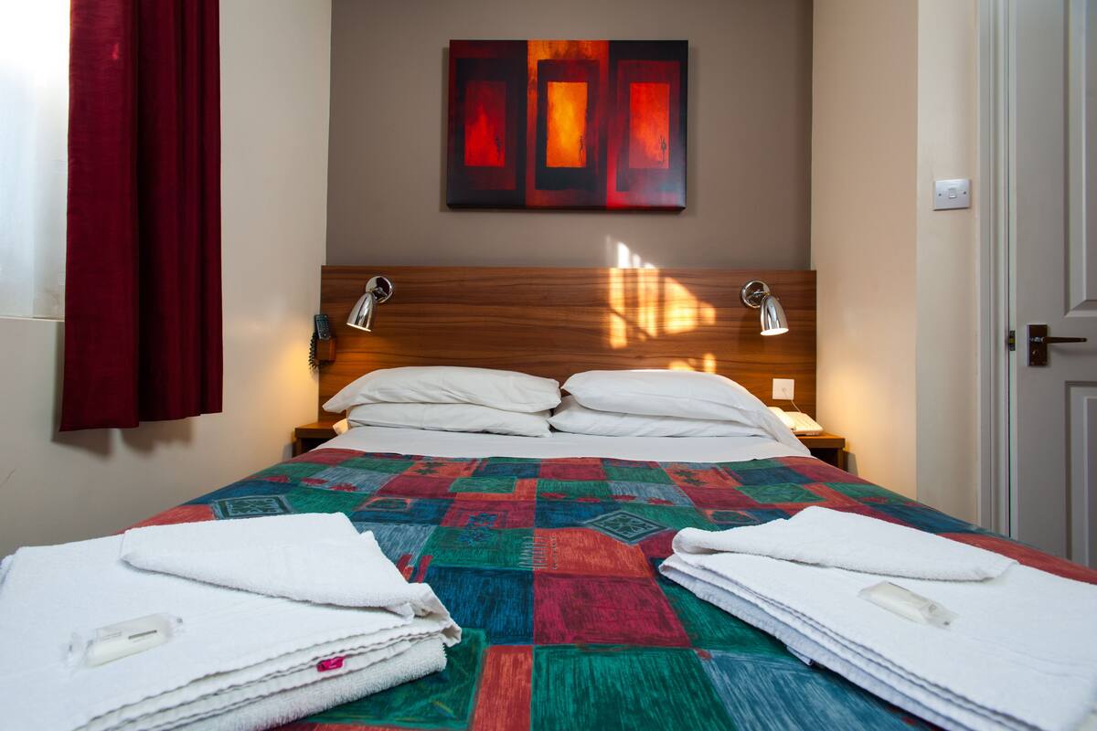Quarto de hotel com cama, duas luminárias de cabeceira, cortina vermelha, uma obra de arte na parede e uma porta.