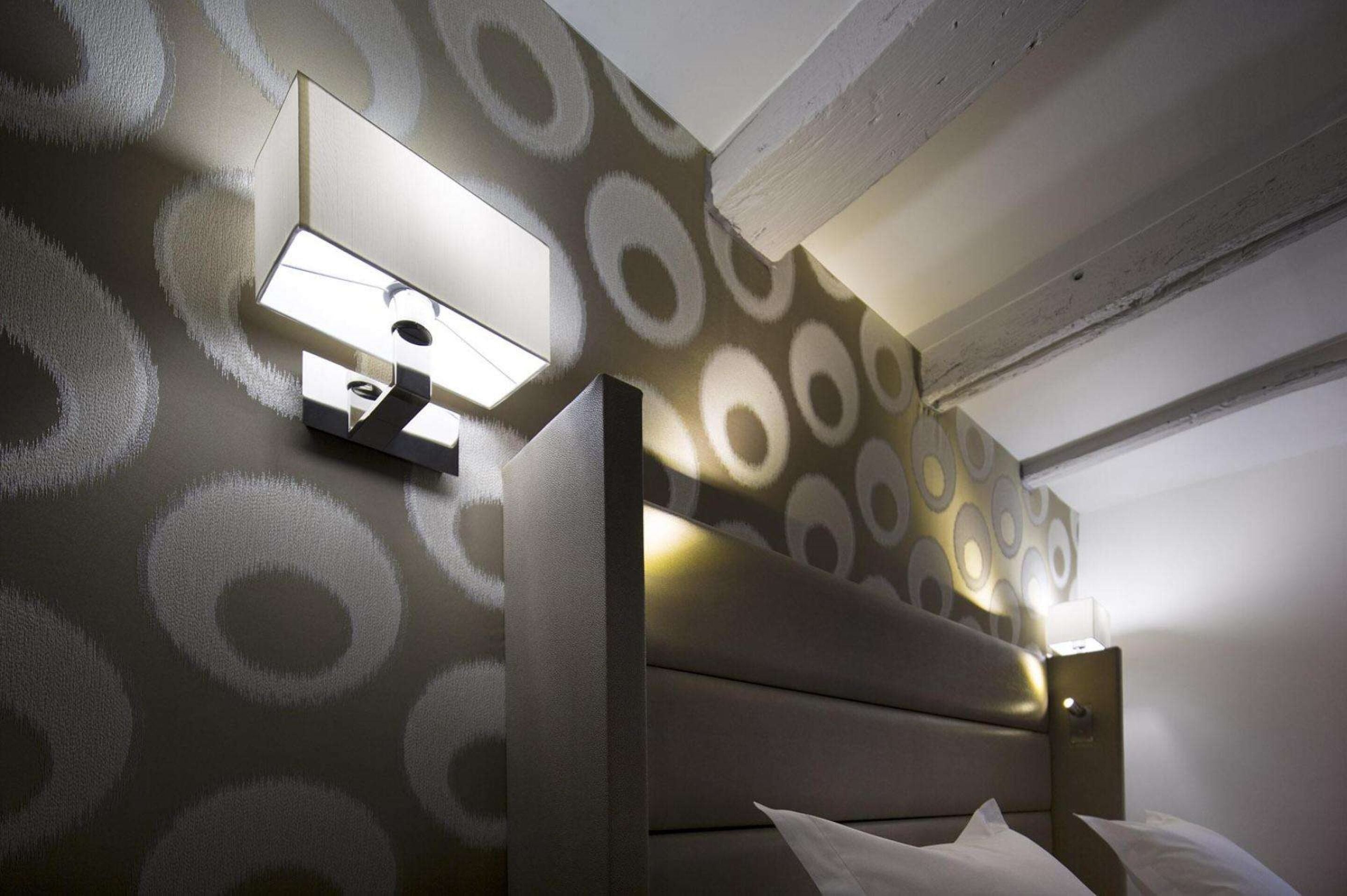 Quarto de hotel com parede estampada, cama com roupa de cama branca e mesa de cabeceira com abajur.