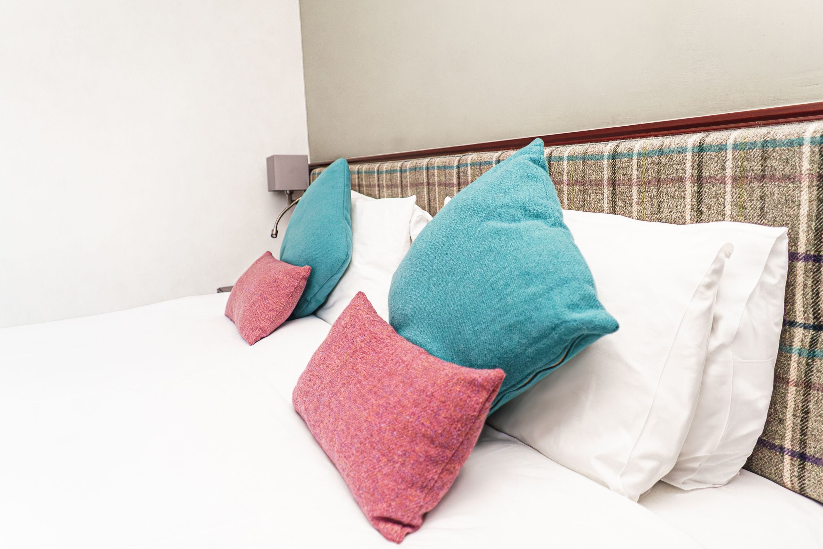 Cama com almofadas brancas e azul-turquesa, cabeceira xadrez e parede lisa.