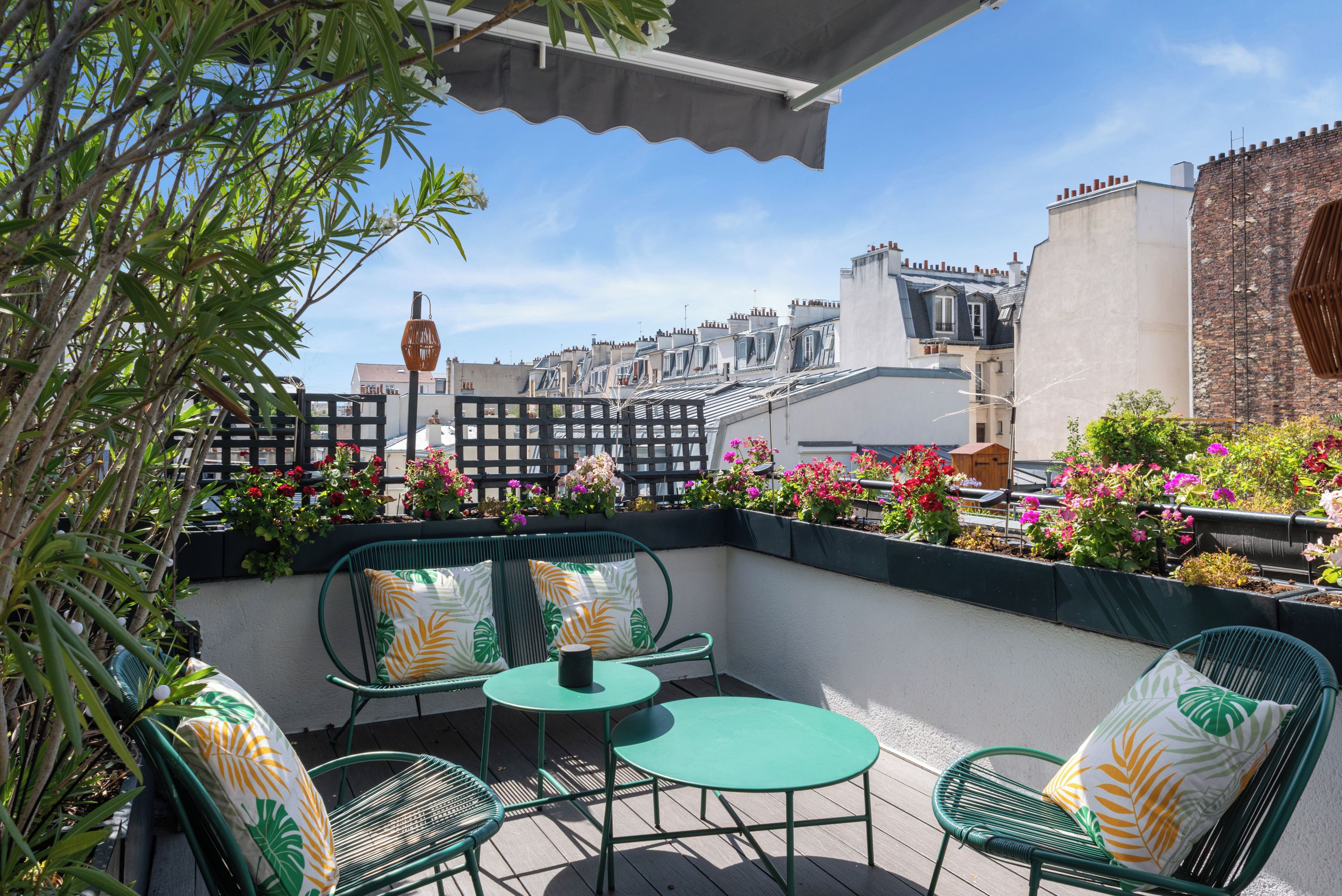 Terraço no telhado com cadeiras e mesa de metal verde, cercado por plantas e flores em vasos.