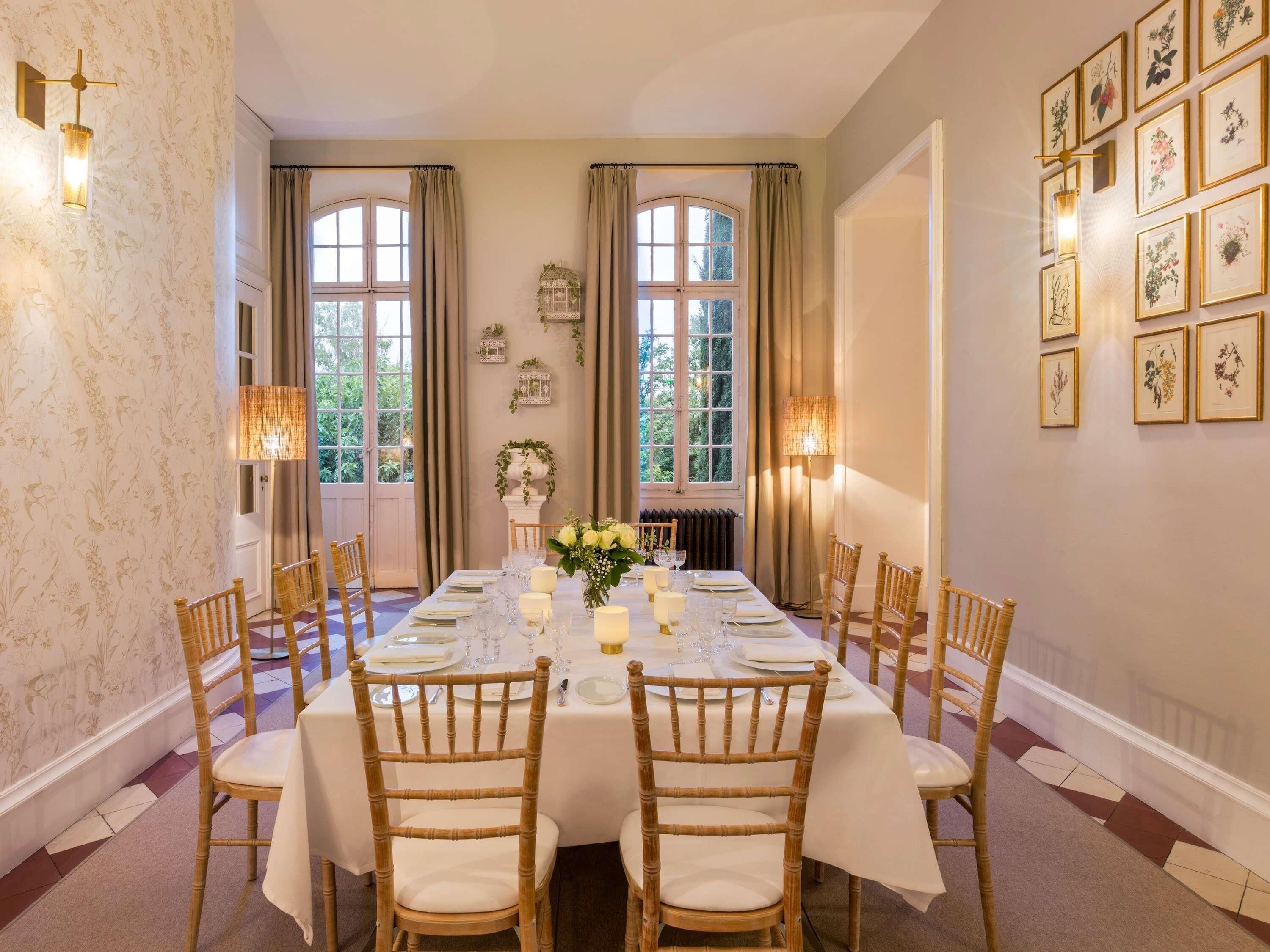 Uma sala de jantar com uma mesa comprida posta para seis pessoas, ladeada por grandes janelas com cortinas, uma parede com impressões botânicas emolduradas e um papel de parede estampado.