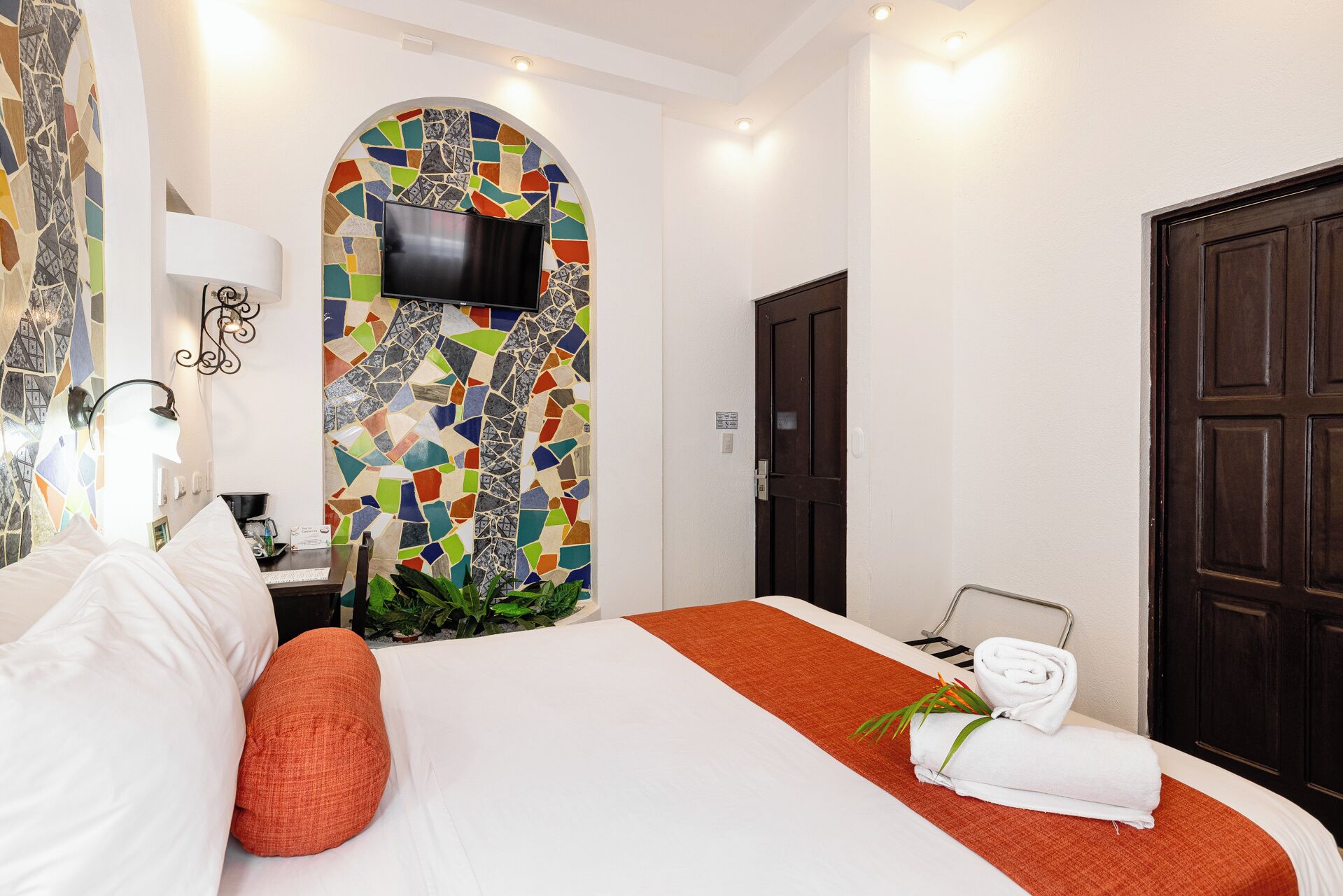 Quarto de hotel com uma cama grande, televisão presa a uma parede com mosaico colorido, porta de madeira escura e uma pequena planta na mesinha de cabeceira.